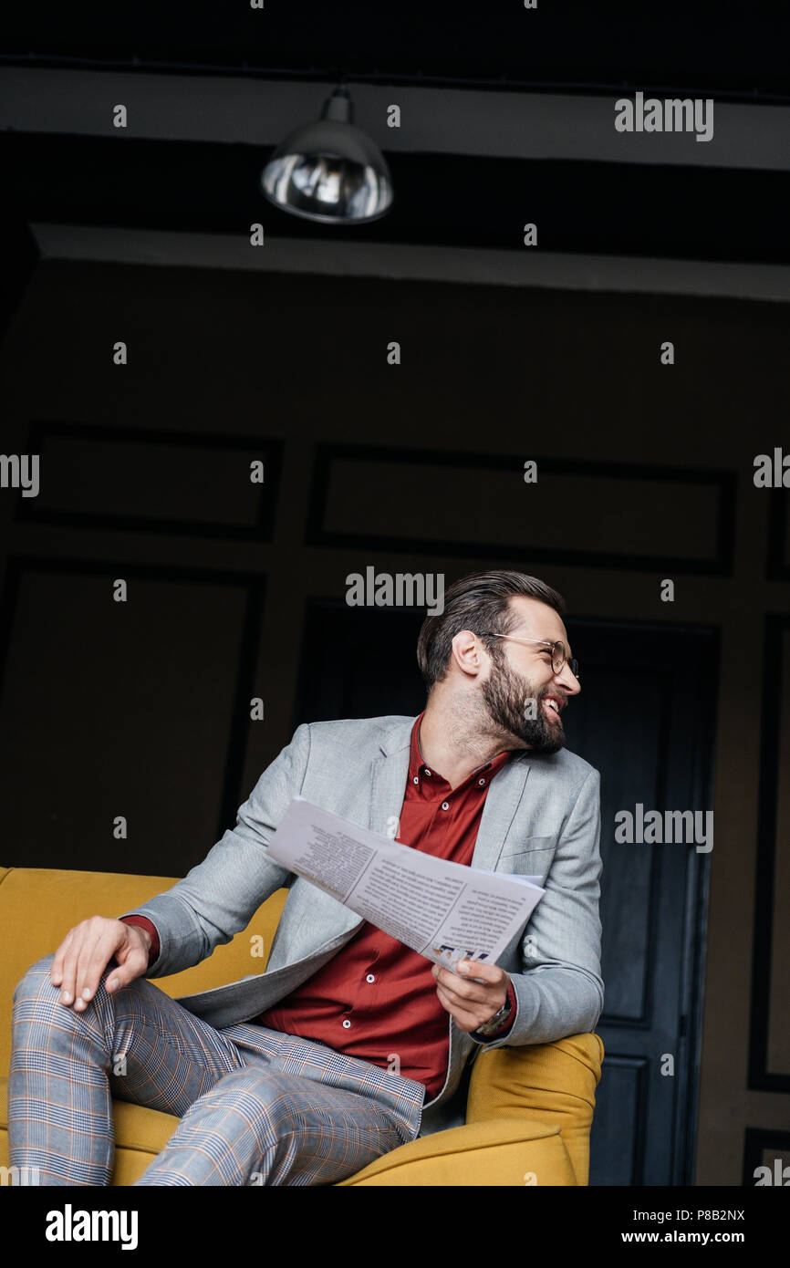 stylish man with newspaper in trendy grey suit sitting on couch in loft ...