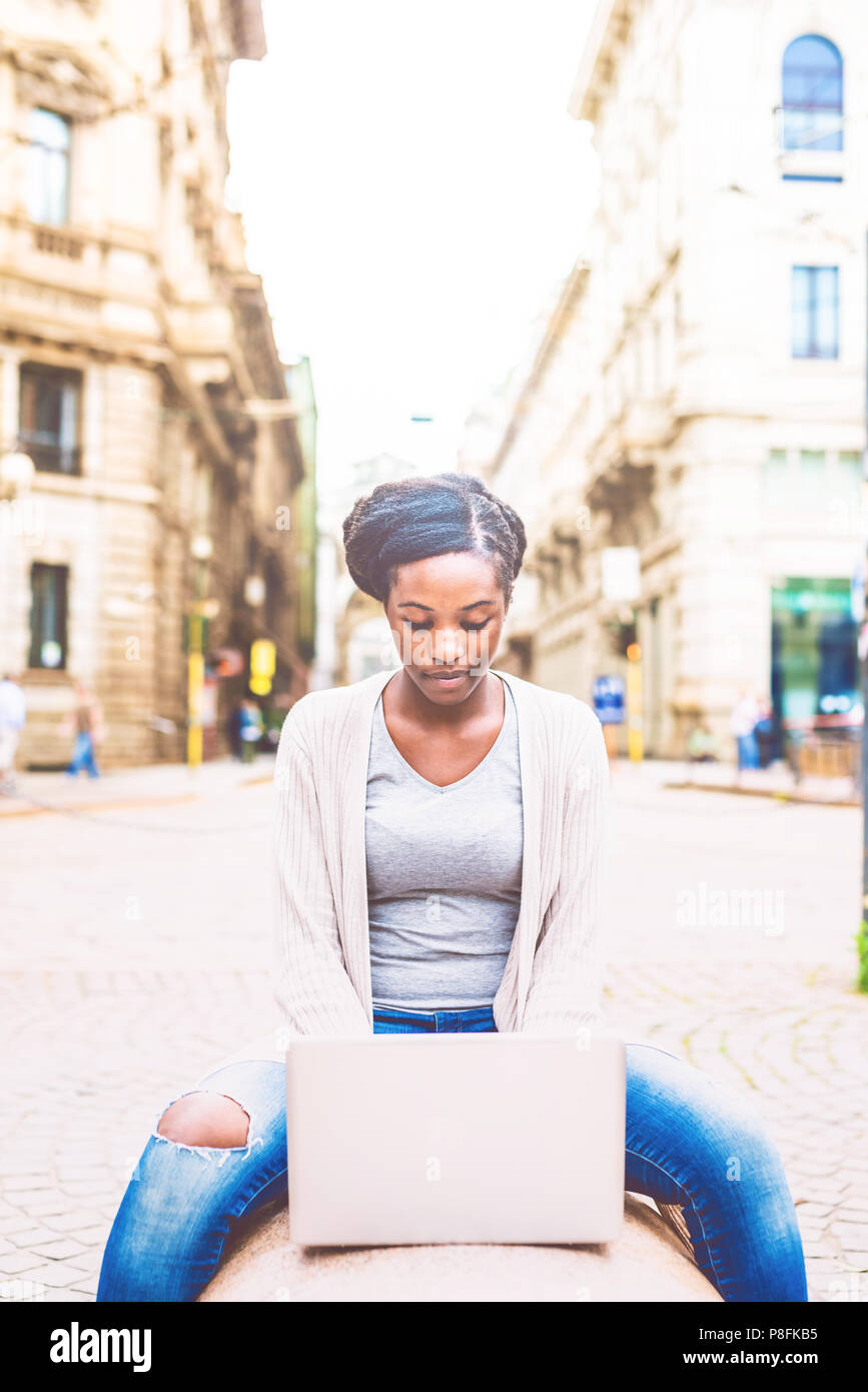 Young black woman using computer outdoor in the city sitting on a bench ...