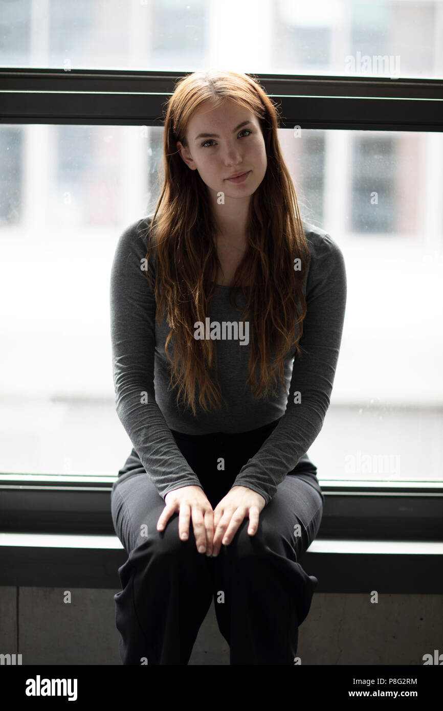 Portrait of a pretty girl sitting on a window ledge with her hands on ...