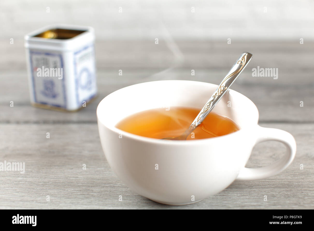 Cup of hot amber tea with steam, with metal tin placed on gray wood ...