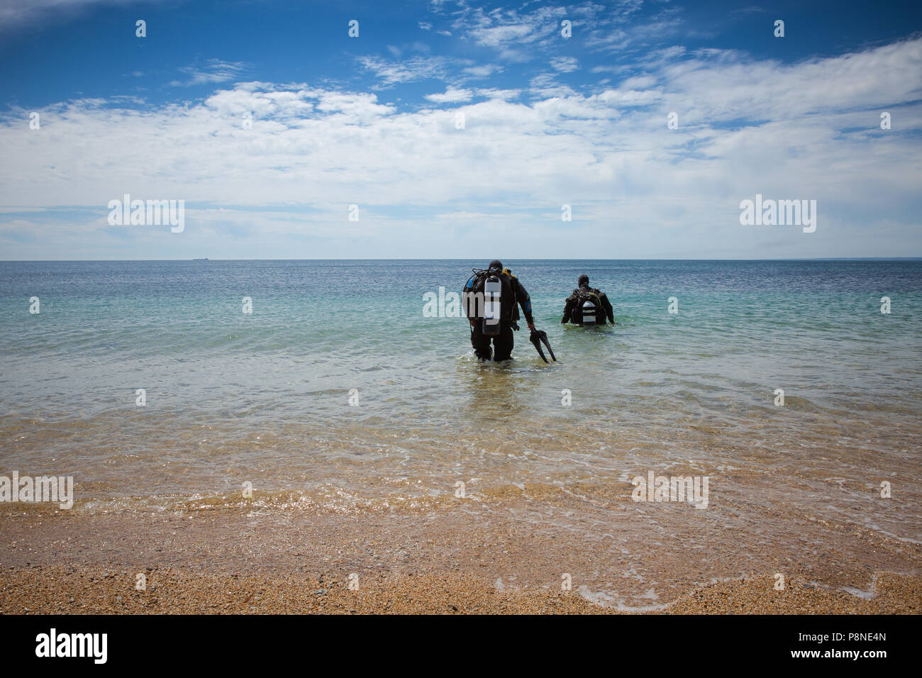 A rear view of two deep sea divers walking into the ocean from a beach ...