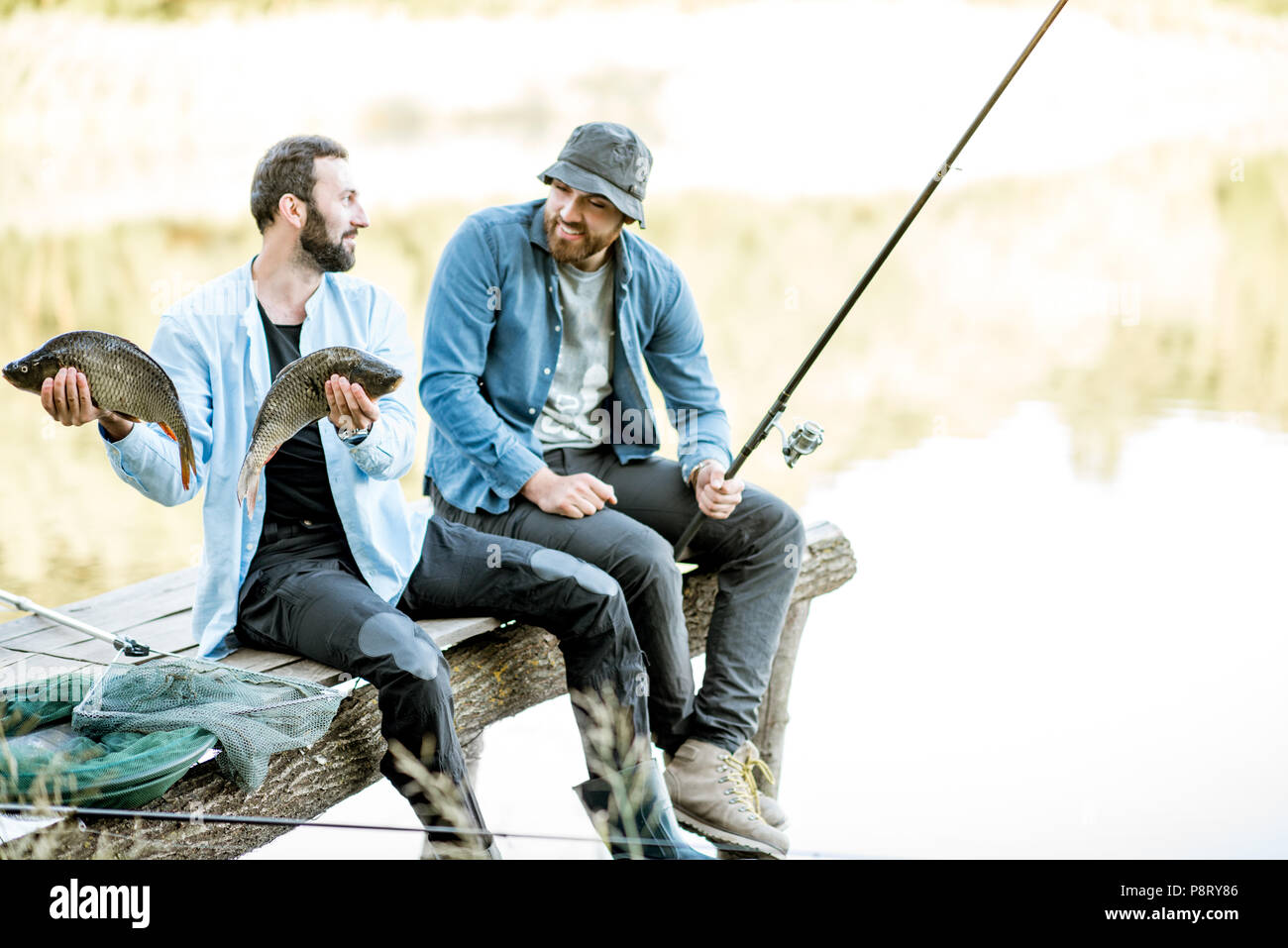 Two happy fishermen holding caught fish sitting on the wooden pier ...