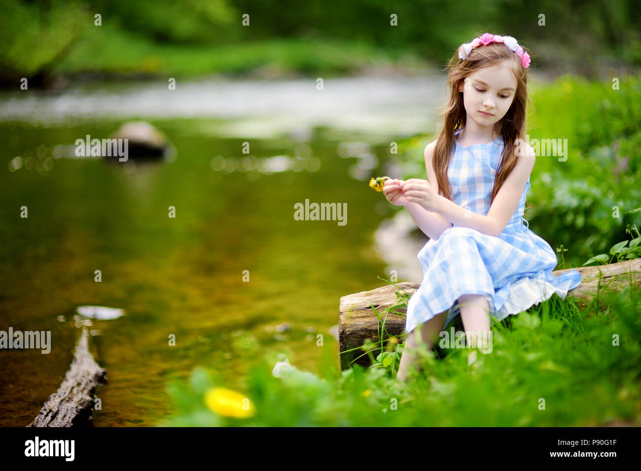 Cute little girl sitting on a log by a river on warm and sunny summer ...