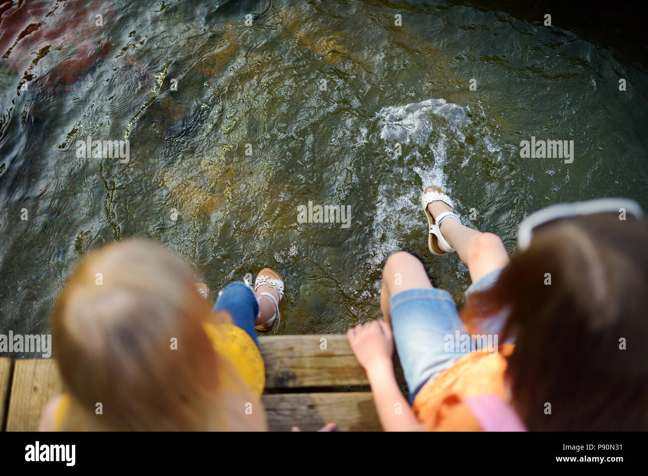 Two cute little girls sitting on a wooden platform by the river or lake ...