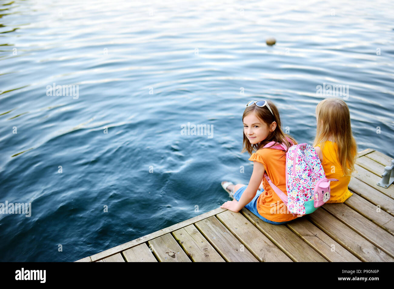 Two cute little girls sitting on a wooden platform by the river or lake ...