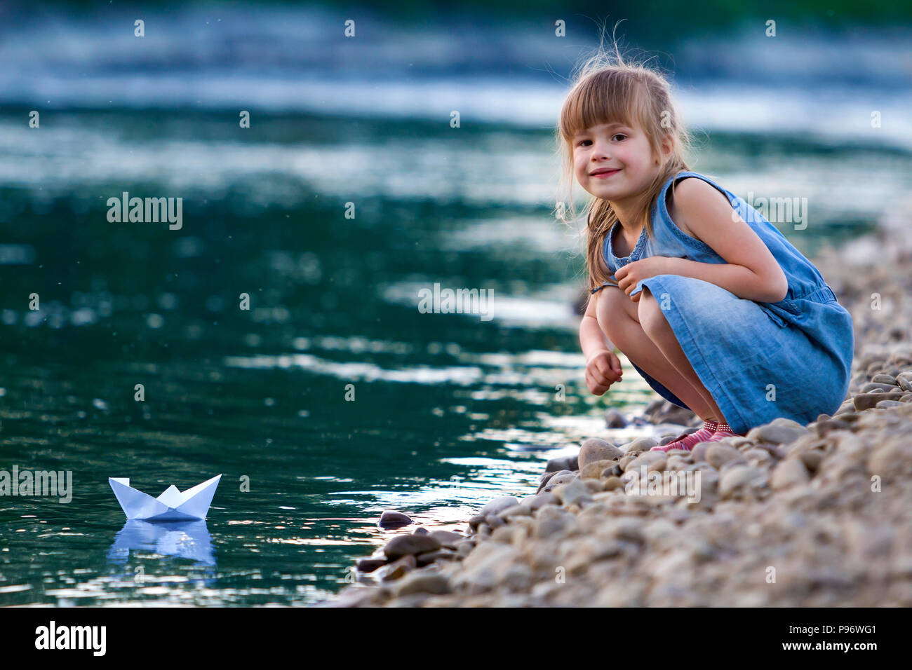 Adorable little cute blond girl in blue dress on riverbank pebbles ...
