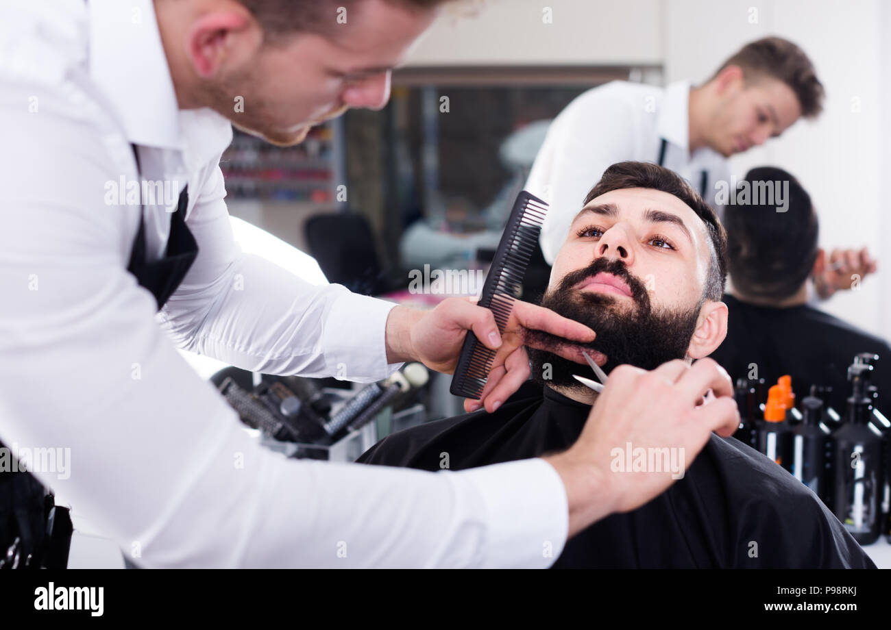 Positive man hairdresser forming beard of client into new shape at hair ...