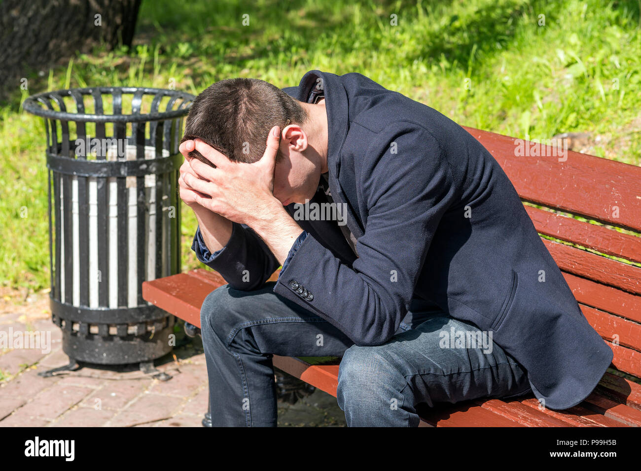 young man sitting on bench in park, covering his face with his hands ...