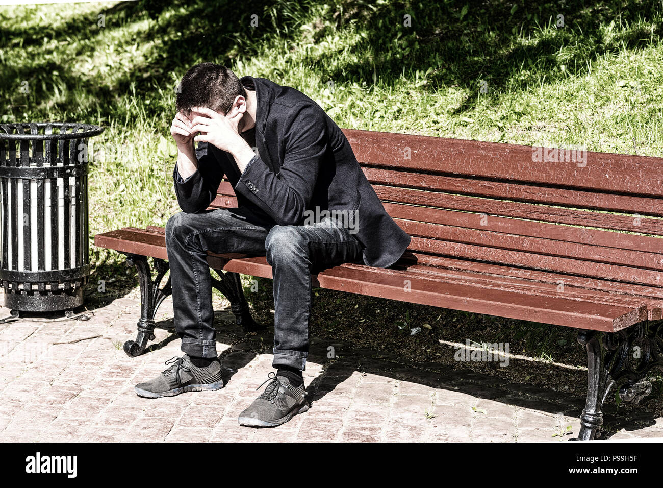 young man sitting on bench in park, covering his face with his hands ...