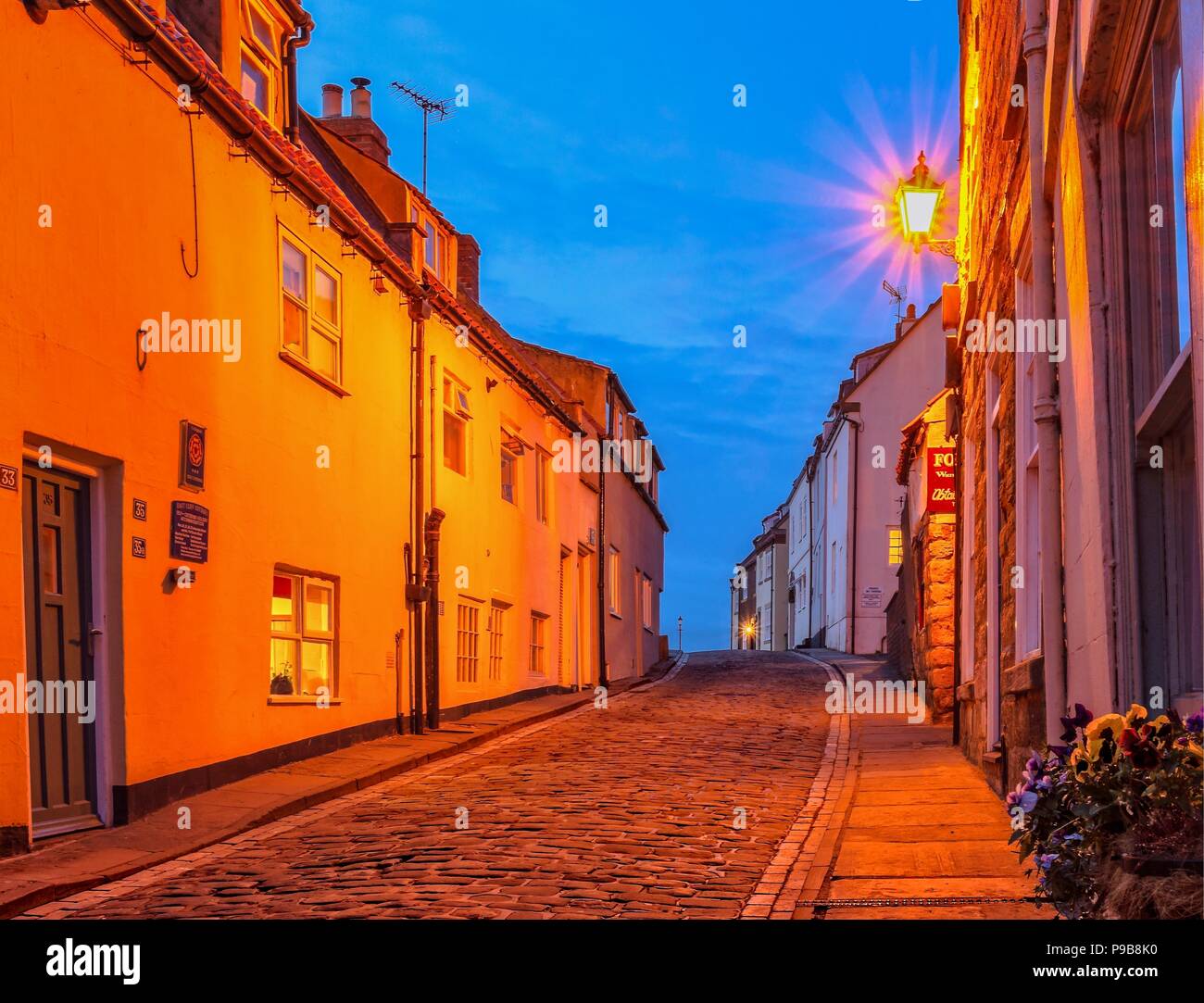 Cobbled street in Whitby at night. Lights shine from a street lamp and ...