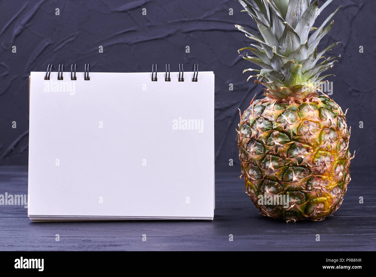 Ripe pineapple and blank paper notebook. Healthy fresh ananas and paper notepad on black ...