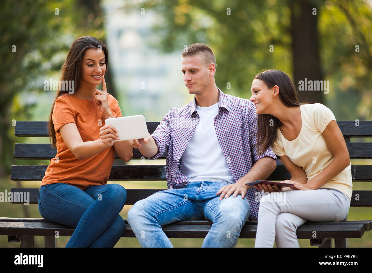 Three friends are sitting on bench in park and talking. They are using ...