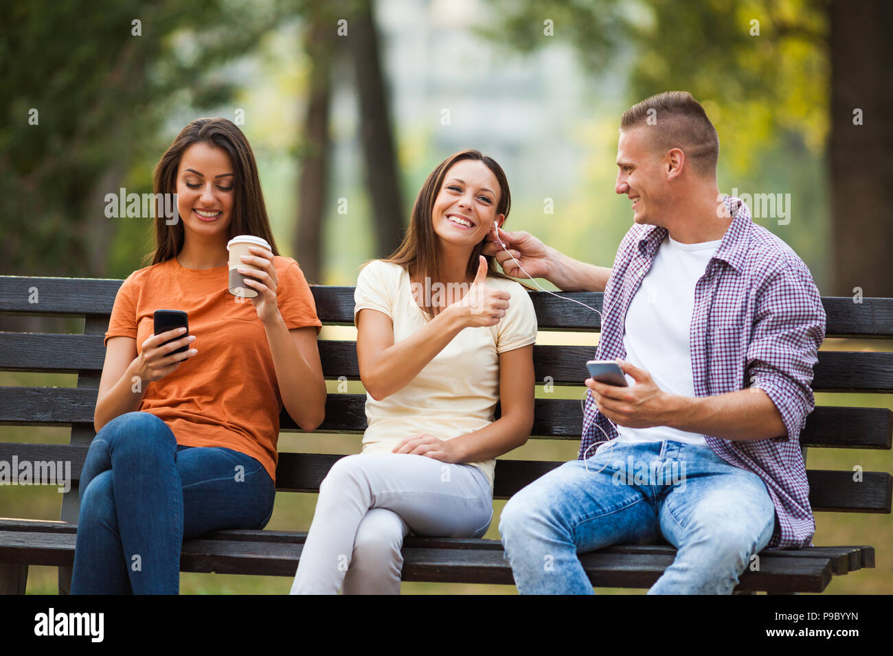 Three friends are sitting on bench in park and using smartphones Stock ...