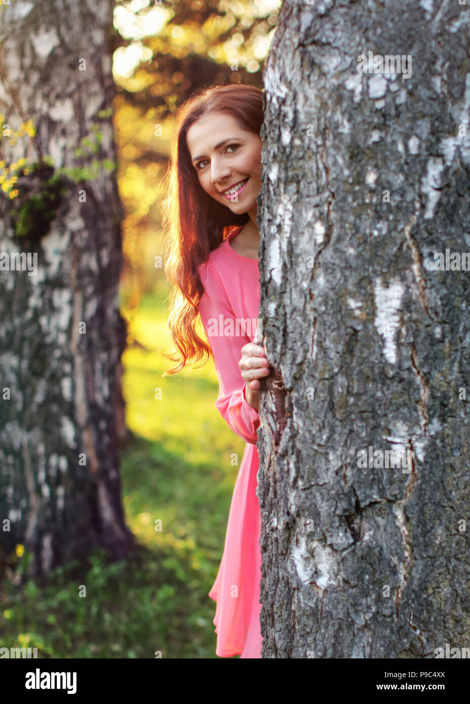 Young woman in pink dress, peek a boo behind the tree, with sunset lit ...