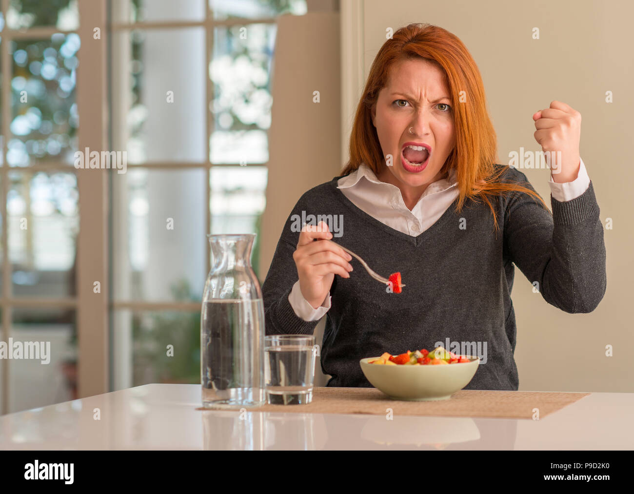 Redhead woman eating fruit bowl, kiwi and strawberry at home annoyed ...