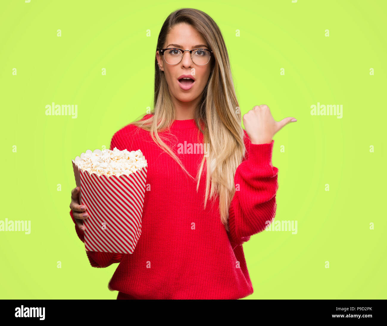 Beautiful young woman eating popcorn pointing with hand and finger up ...