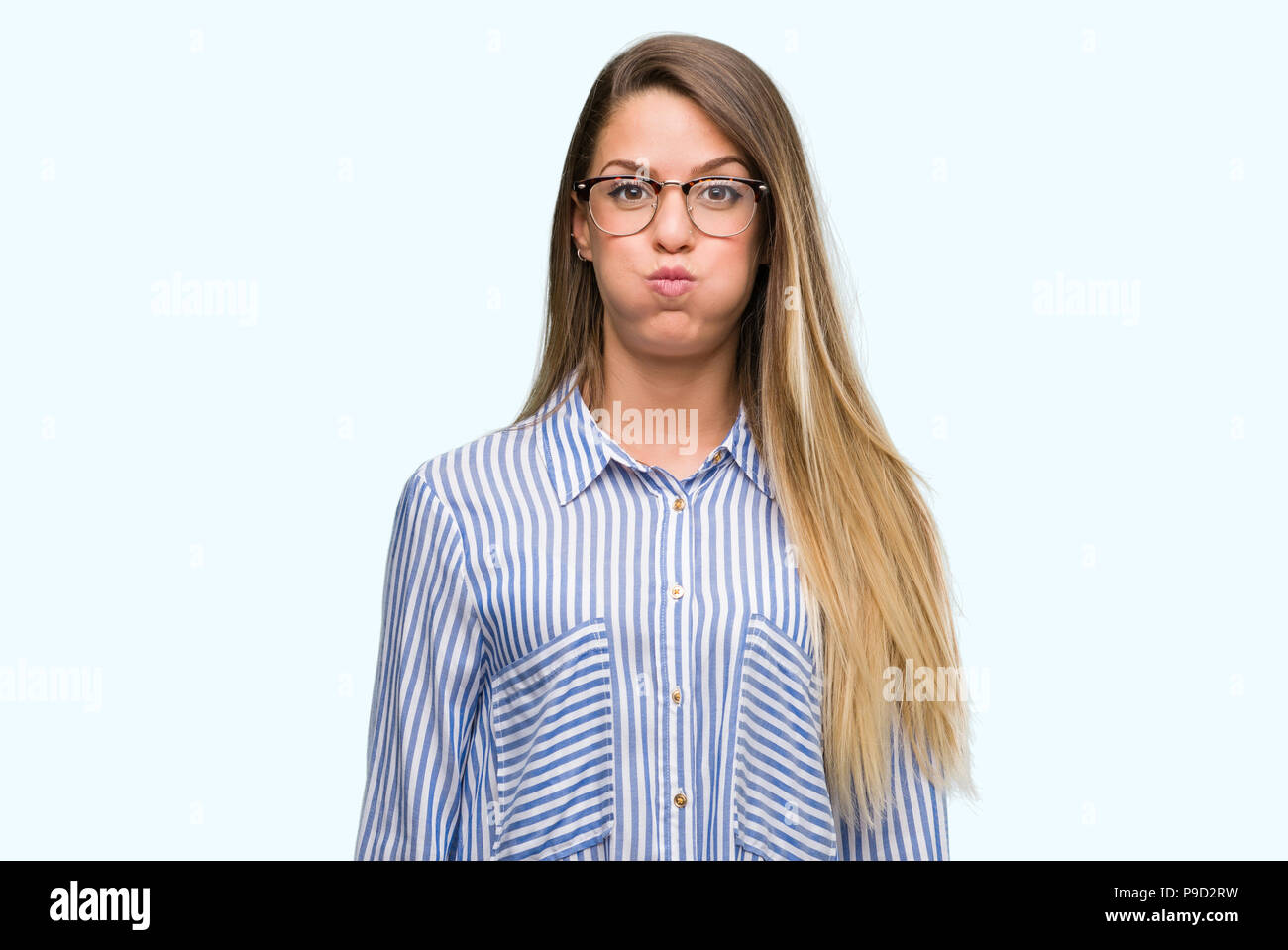 Beautiful young woman wearing elegant shirt and glasses puffing cheeks ...