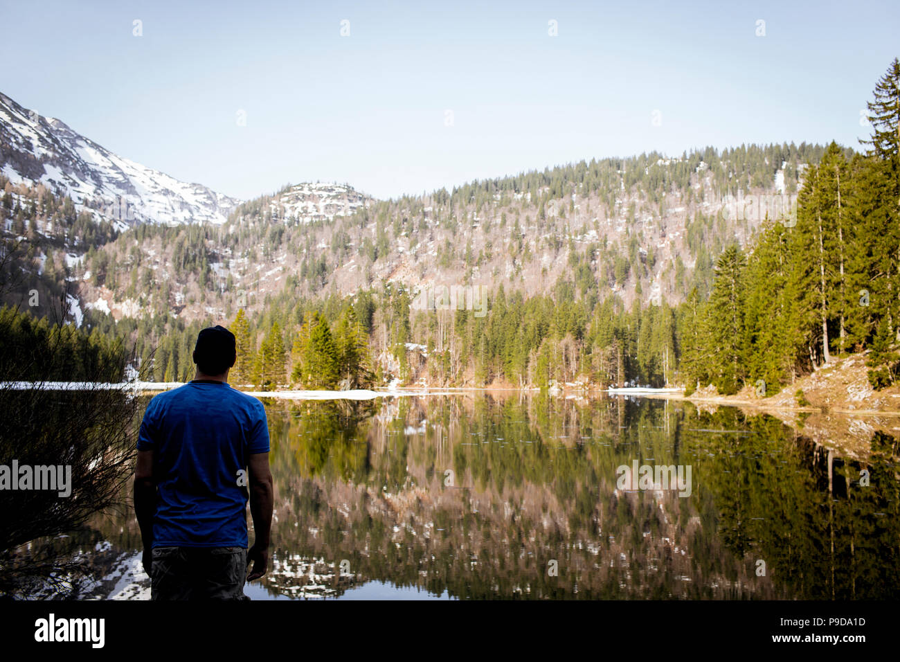 young man with cap and blue shirt is standing in front of mountain lake ...