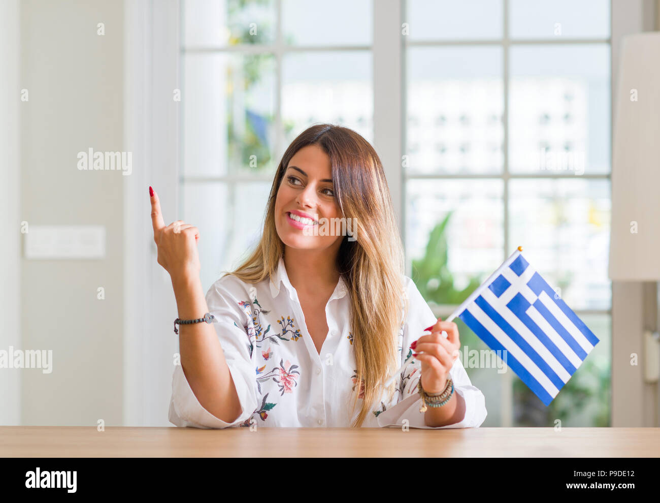 Young woman at home holding flag of Greece very happy pointing with ...