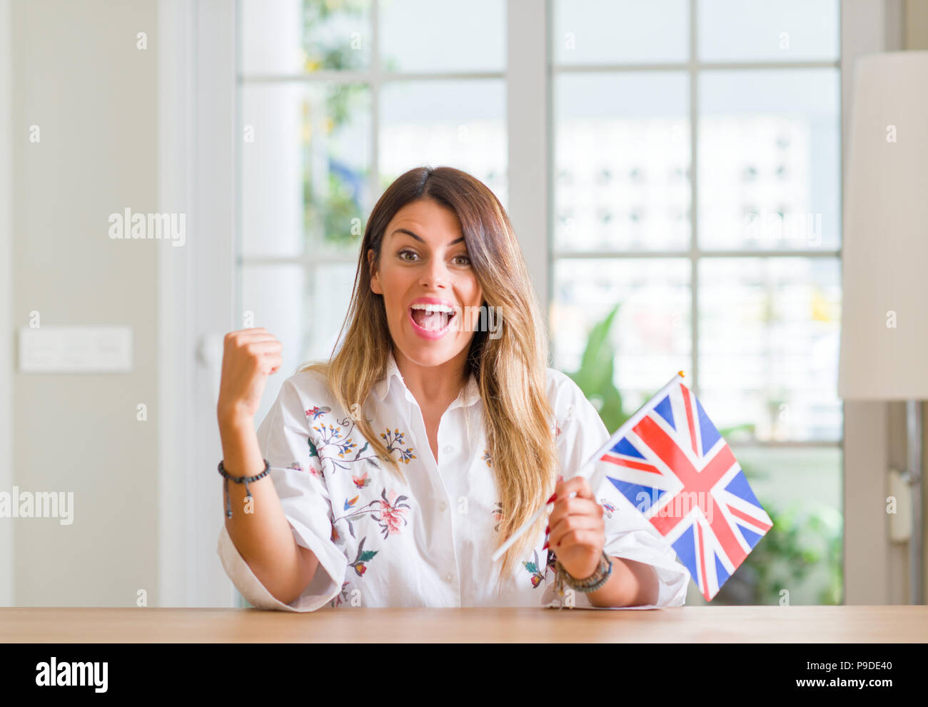 Young woman at home holding flag of United Kingdom screaming proud and ...