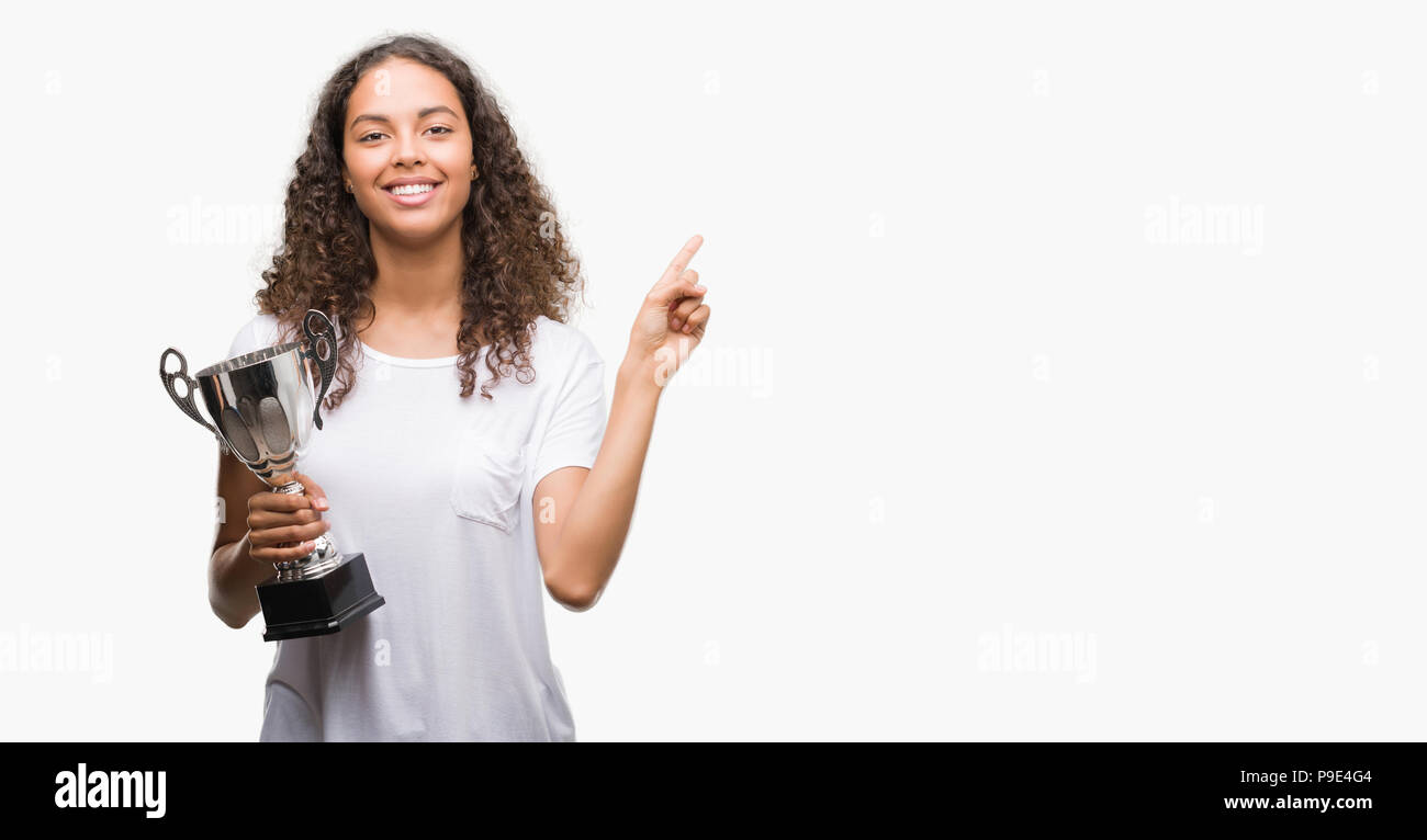 Young hispanic woman holding trophy very happy pointing with hand and ...