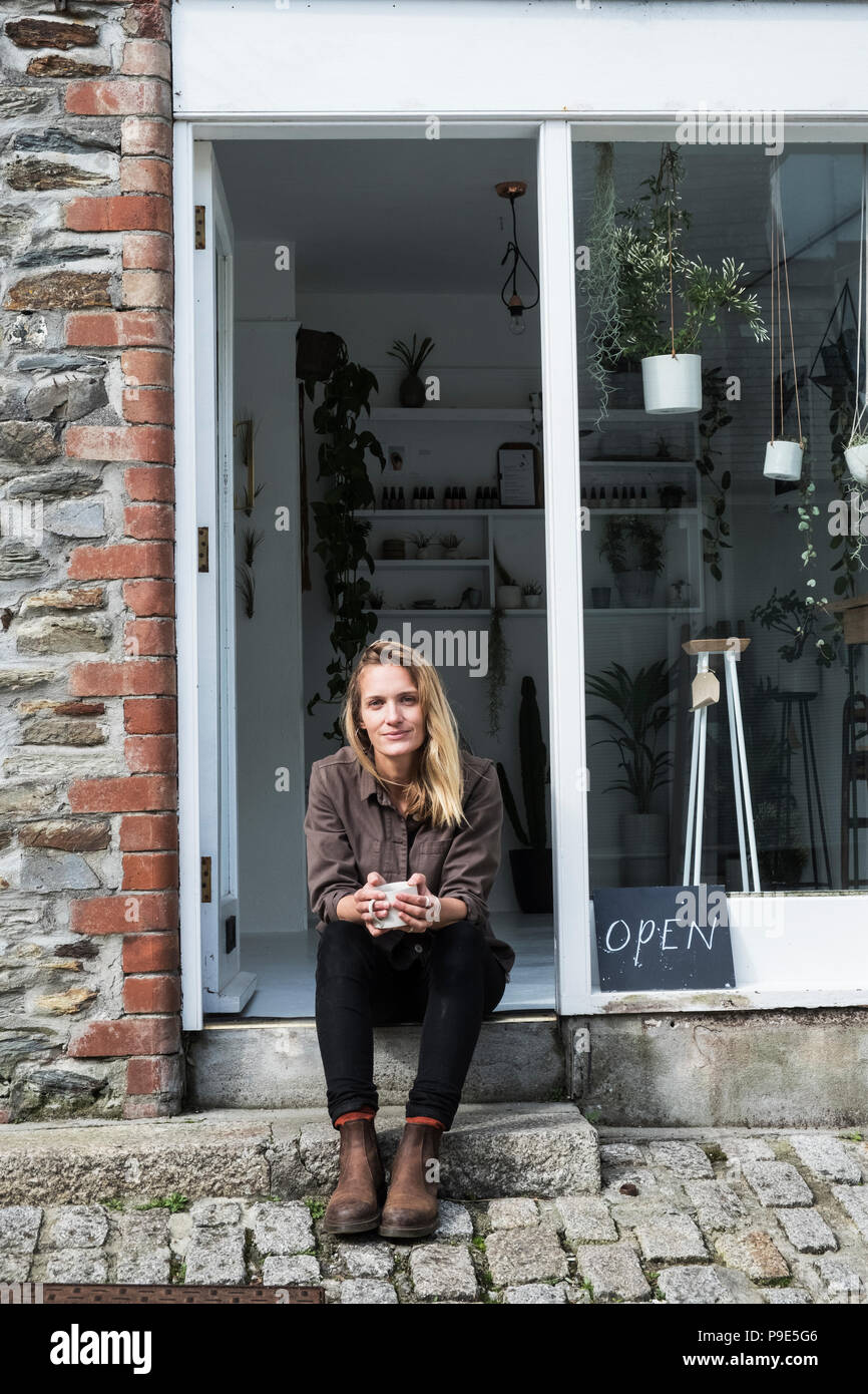 Smiling female owner of plant shop sitting on steps outside her store ...