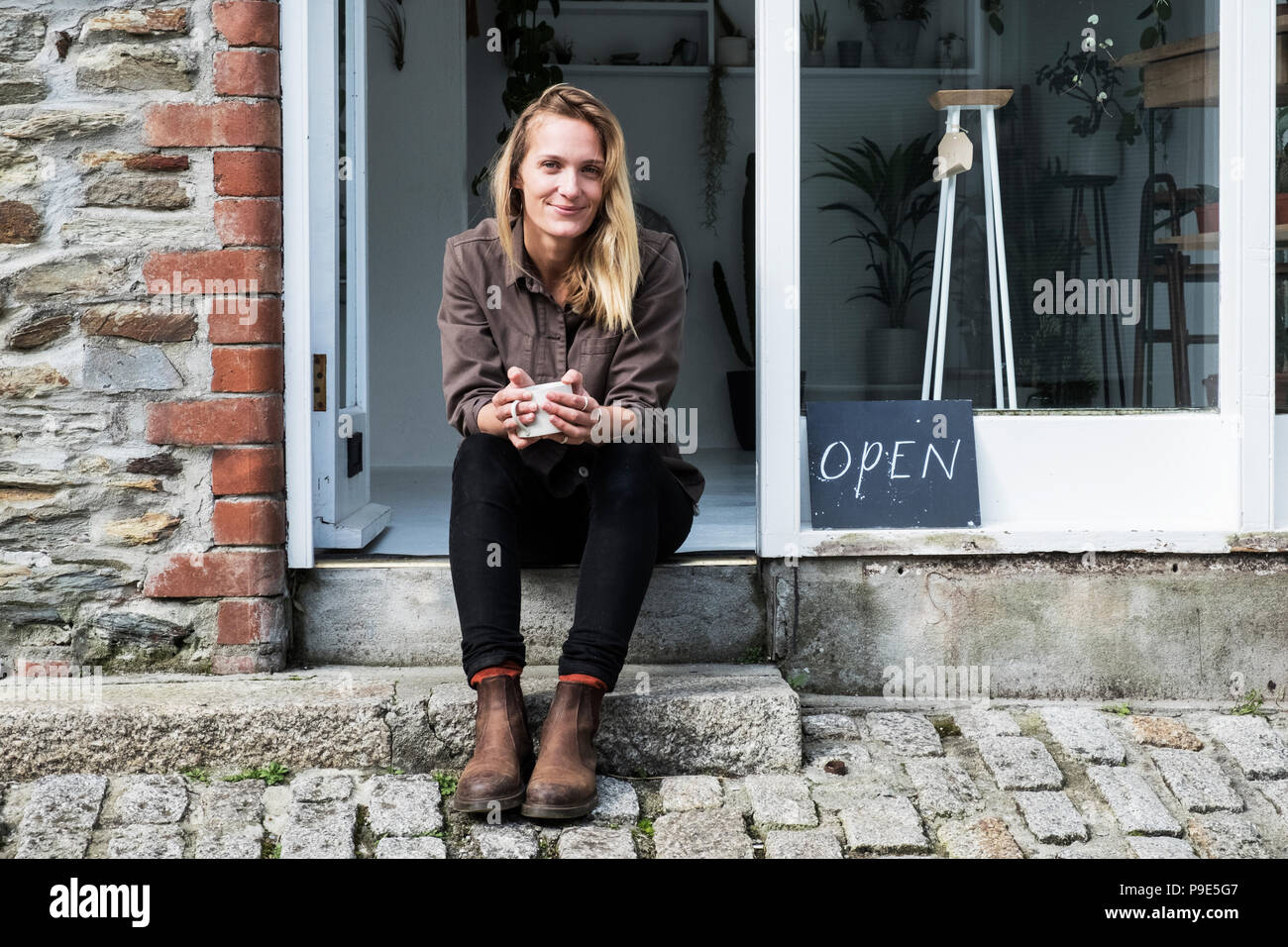 Smiling female owner of plant shop sitting on steps outside her store ...