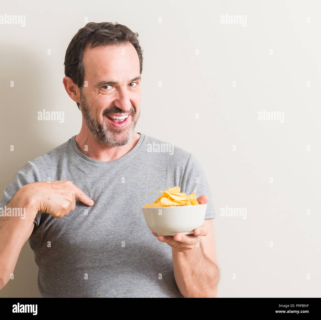 Senior man eating potato chips with surprise face pointing finger to ...