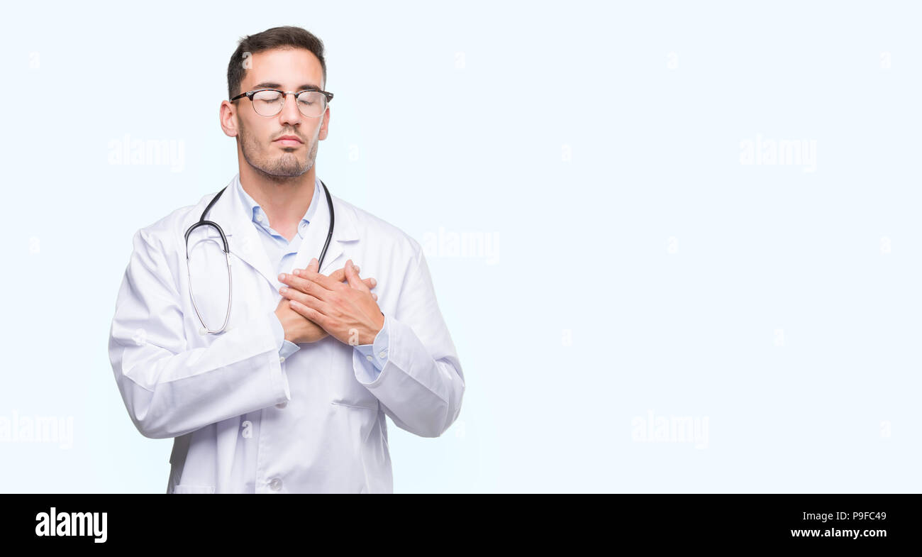 Handsome young doctor man smiling with hands on chest with closed eyes ...