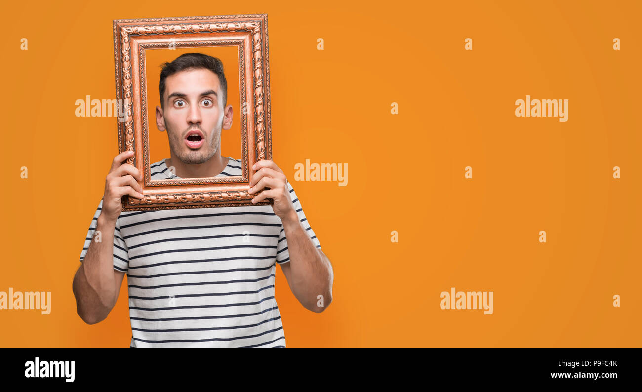 Handsome young man looking through vintage art frame scared in shock ...