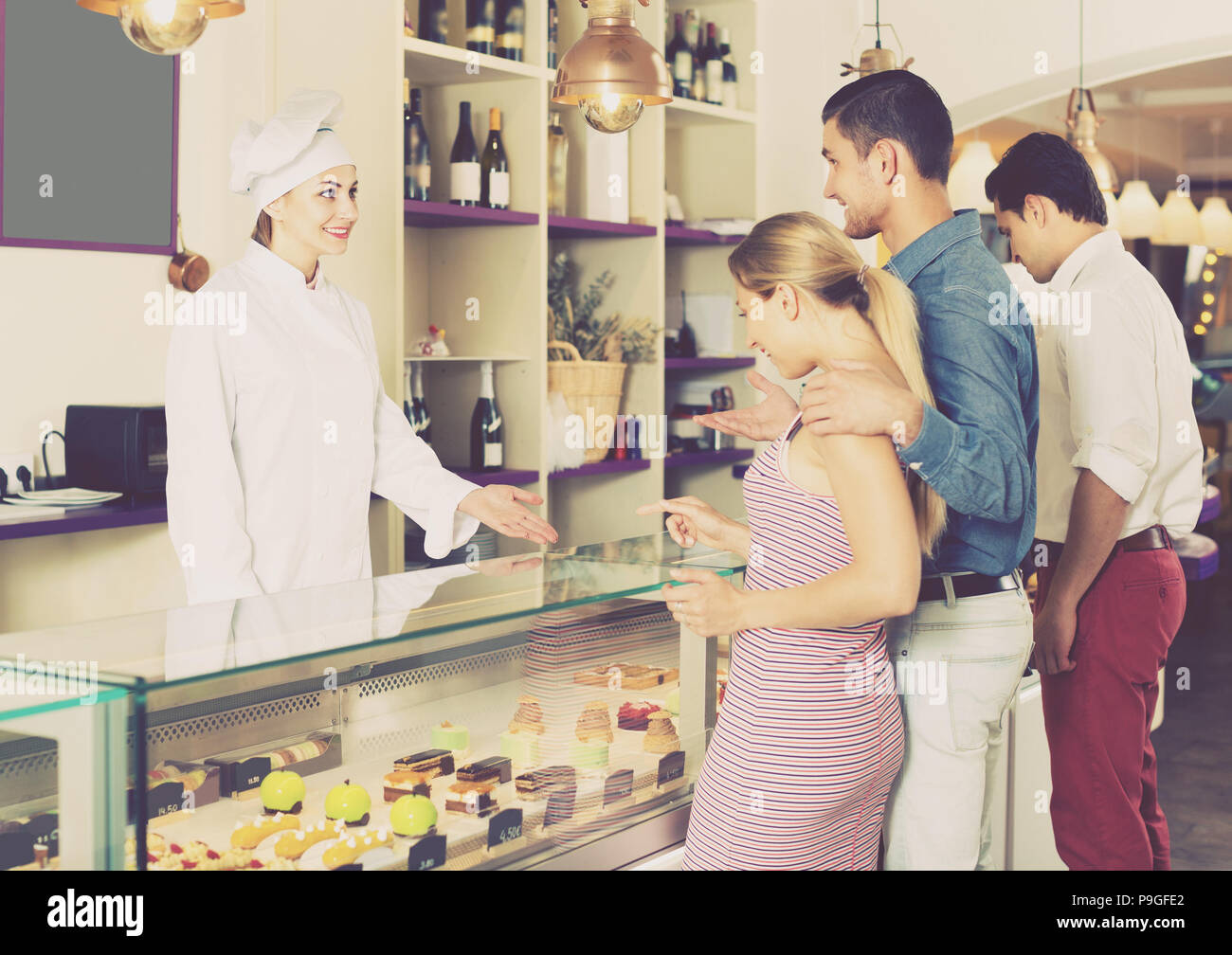 Cheerful pastry chef female is offering cakes from showcase in pastry ...