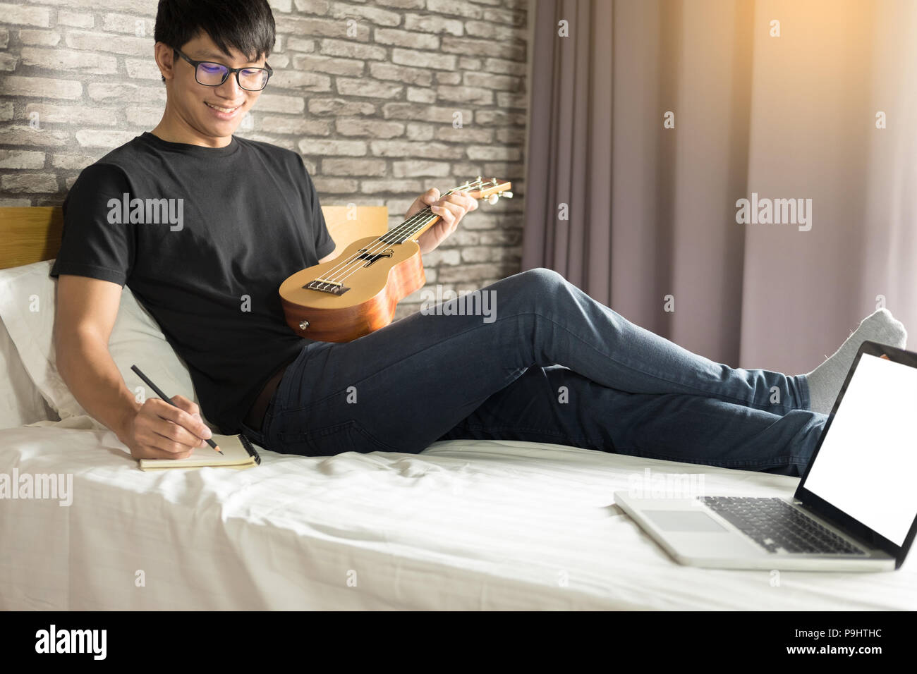 Happy Young Asian Man Playing Ukulele Sitting On Bed In Bedroom Stock