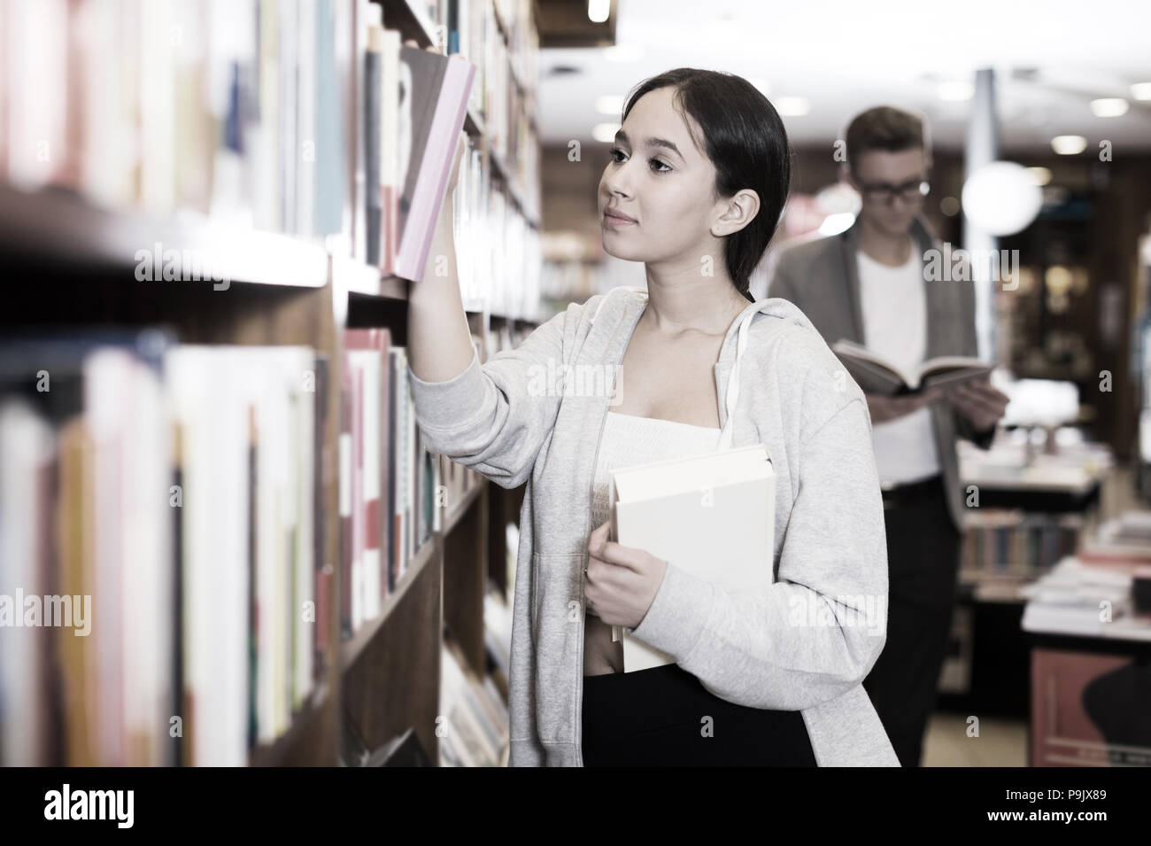 Young positive woman searching for textbooks on bookshelves in shop ...