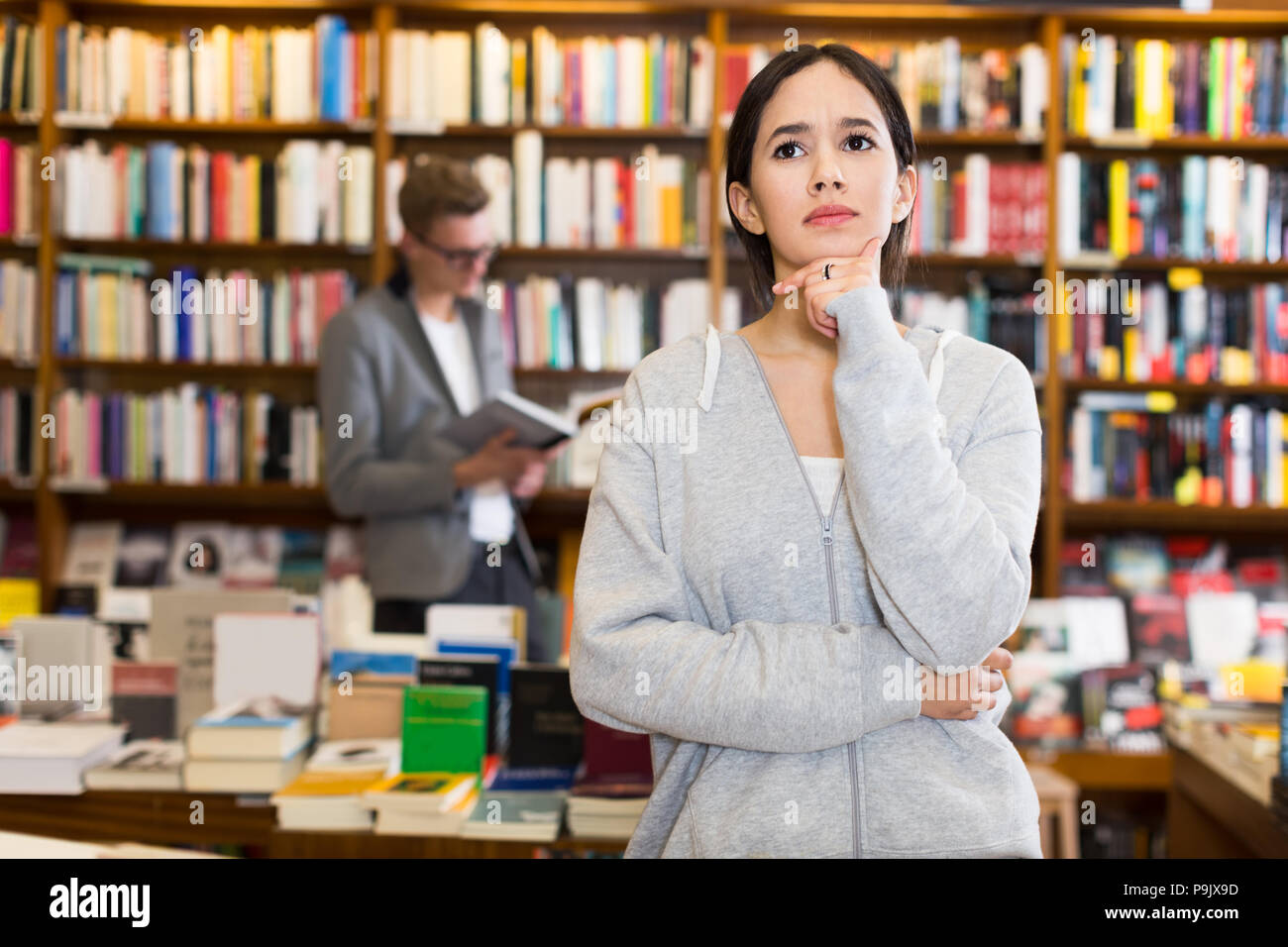 Thoughtful female student standing in bookshop on background with ...