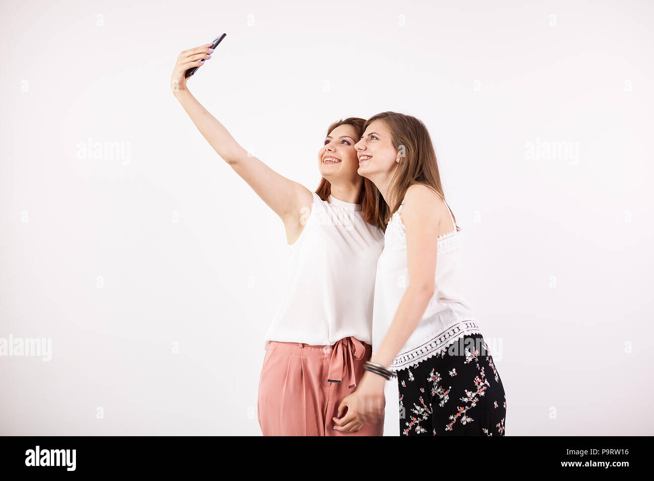 Two happy gorgeous young woman friends smiling and taking a selfie over ...