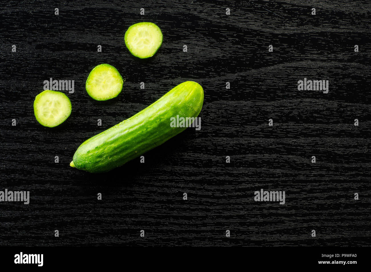 One fresh mini cucumber and three round slices flatlay on black wood ...