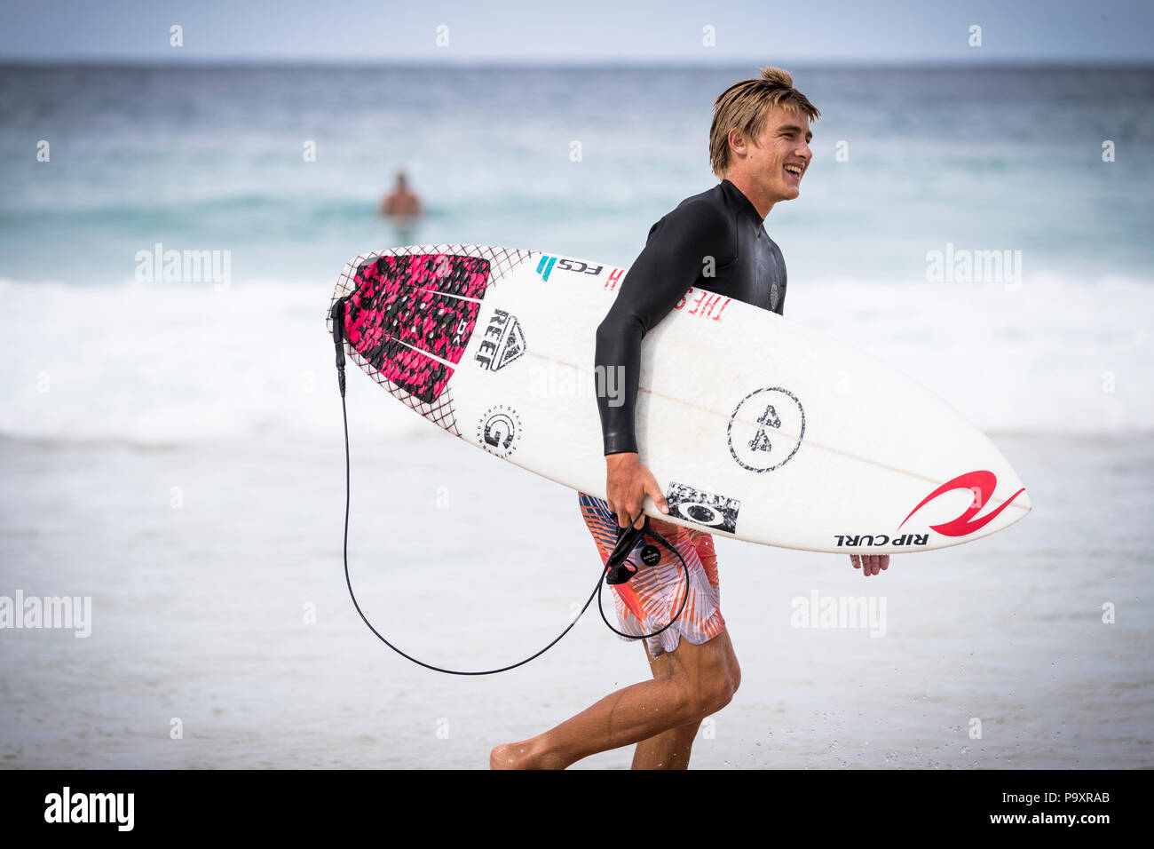Side view of single smiling male surfer carrying surfboard along beach ...