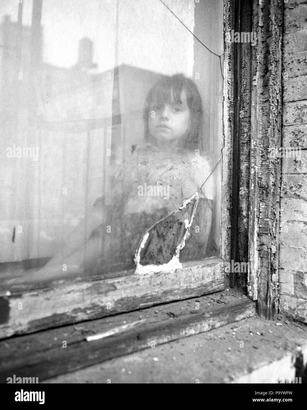 1970s POOR LITTLE GIRL LOOKING AT CAMERA OUT OF BROKEN WINDOW OF SLUM ...