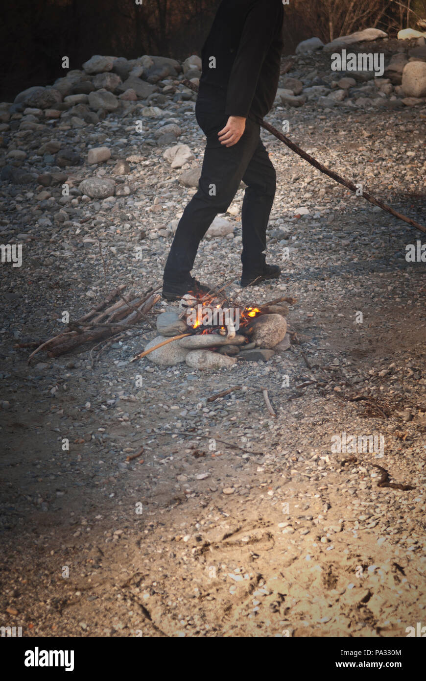 cropped view of adult male tending to an outdoor fire on a pebble beach ...