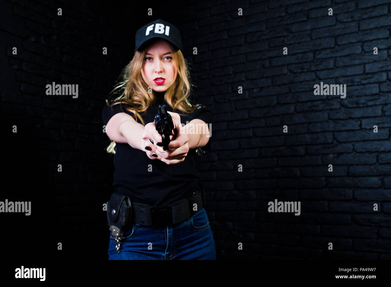 FBI female agent in cap and with gun at studio against dark brick wall ...