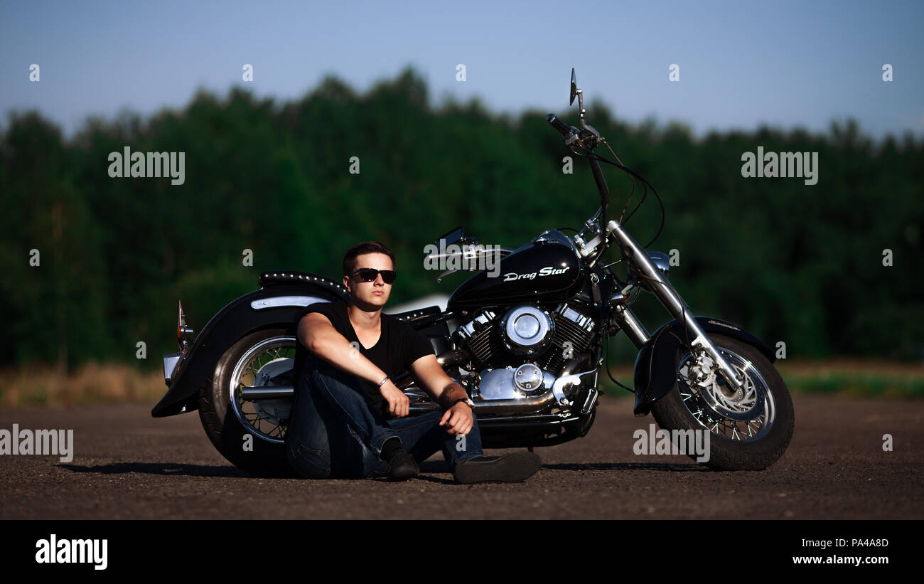 Handsome biker man in glasses sitting at his motorcycle. Countryside ...