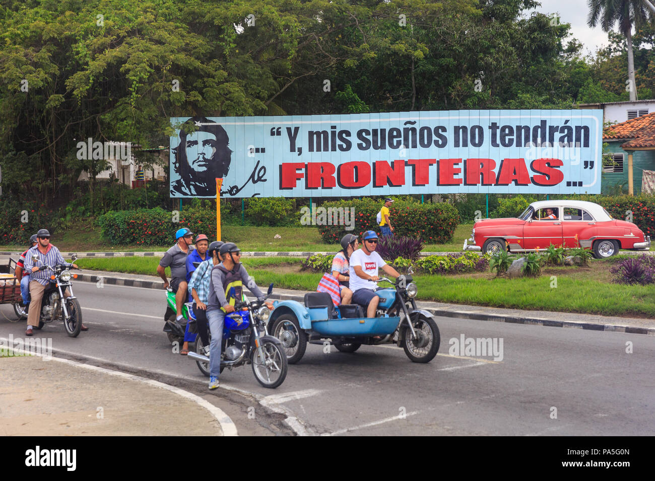 Cuban people in cars and motorcycles in front of propaganda billboard ...