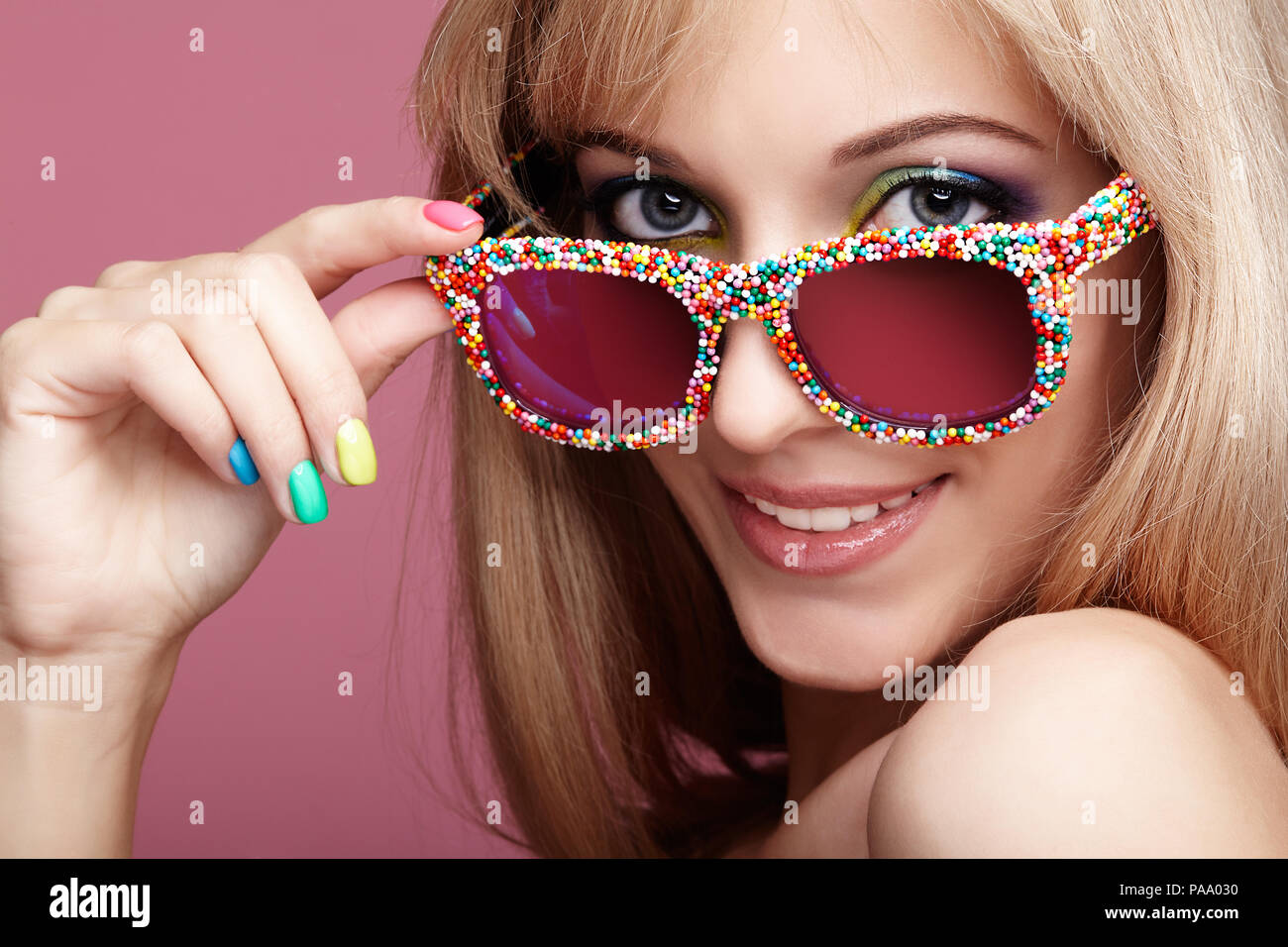 Young blonde smiling woman with fun candy glasses on pink background ...