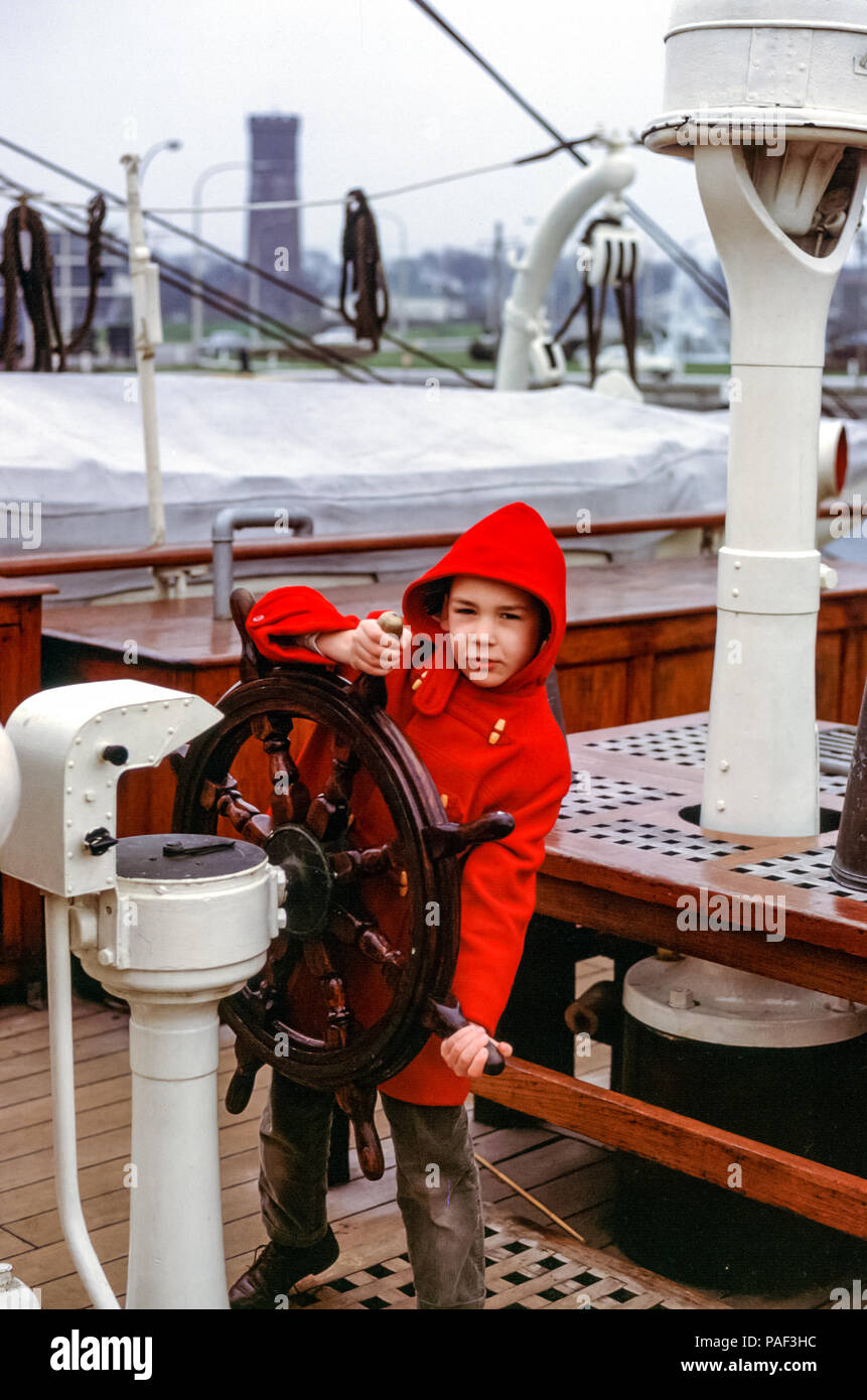 Young boy wearing a red duffel coat steering a ship's captains wheel on ...