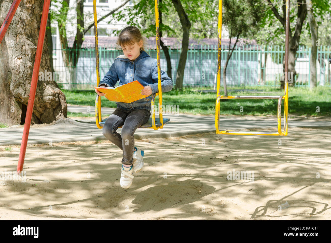 A girl on a walk reading a book swinging on a swing at the playground ...