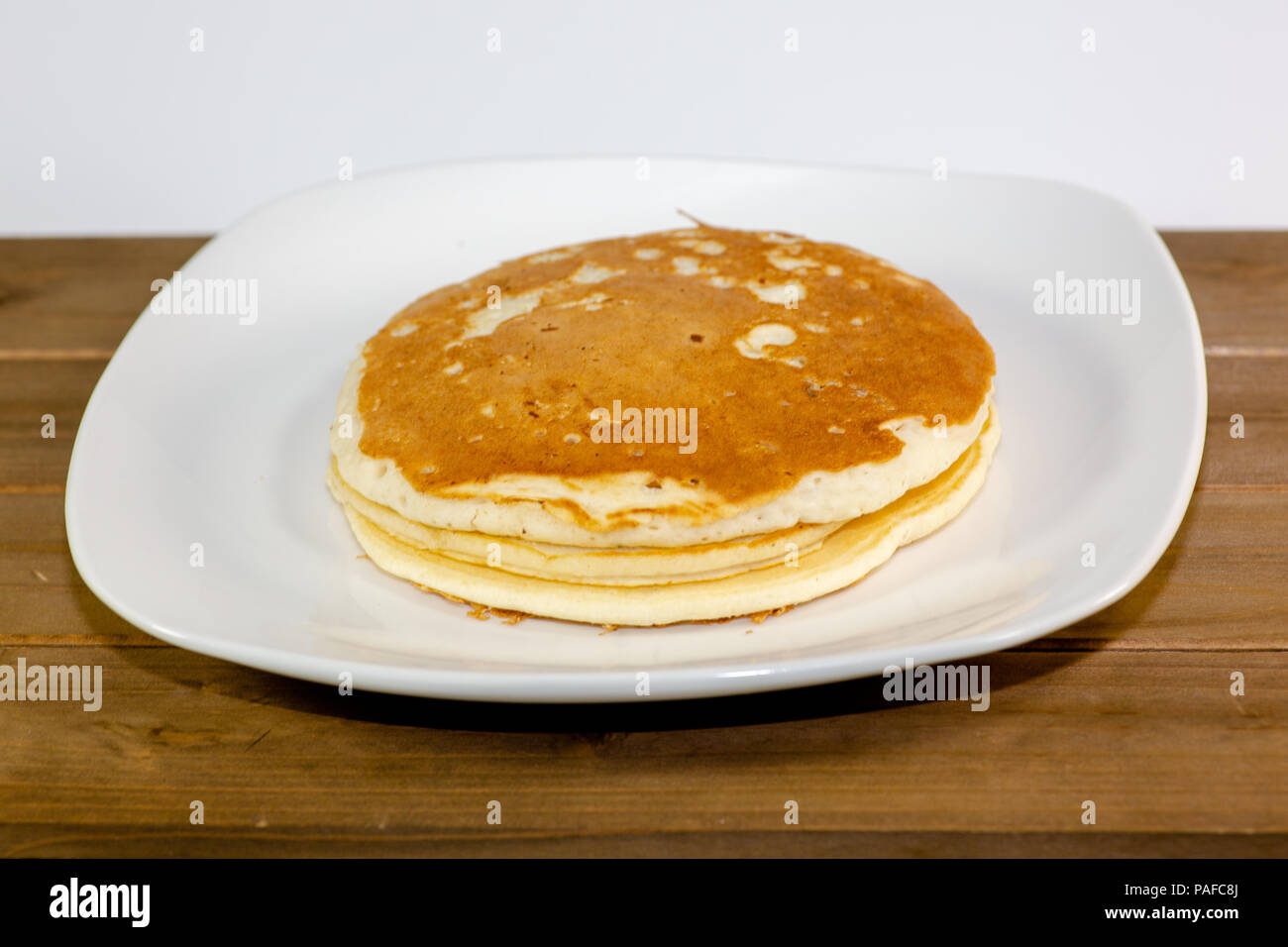 A stack of golden pancakes on a white plate sitting on a kitchen table ...