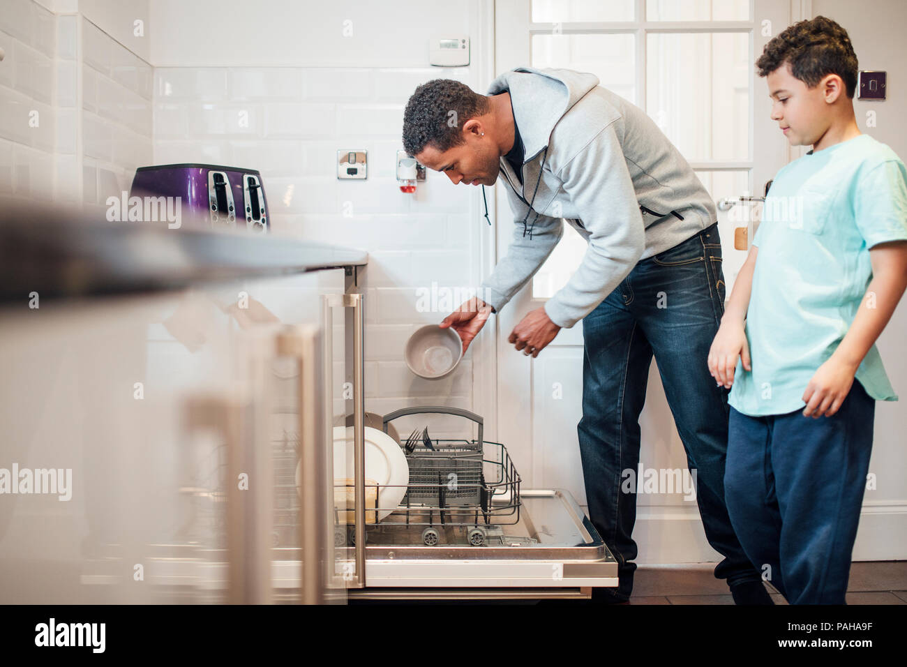 Mid adult man is loading the dishwasher at home and his son is watching ...