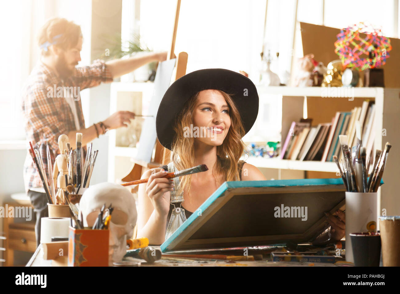 Female long hair artist in cute balck hat smiling and holding brush and ...