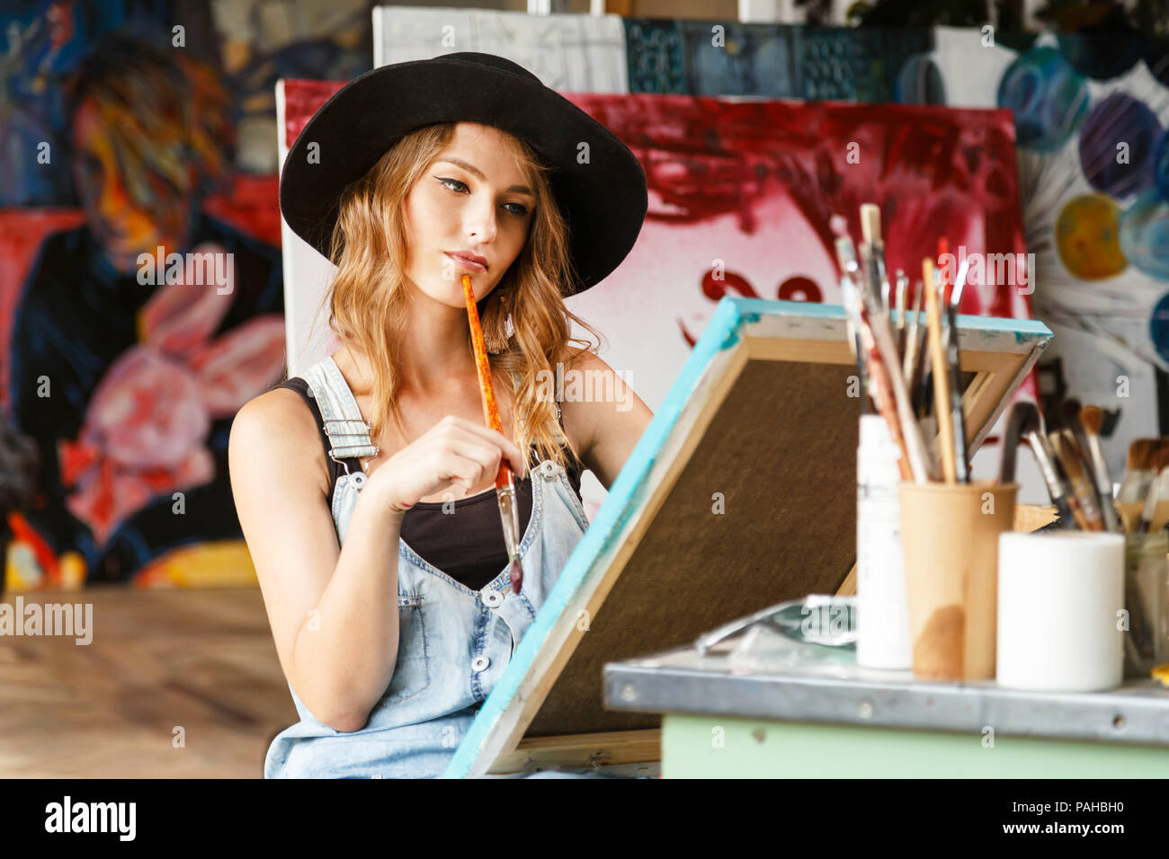 Handsome long-hair female artist in stylish black hat holding canvas ...