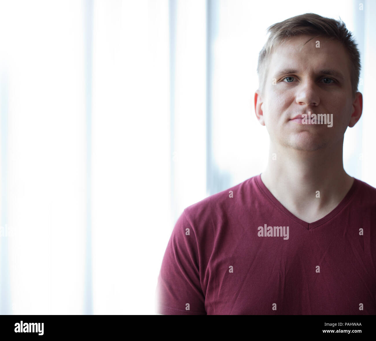 A young man seriously looks at the camera standing near the window in ...